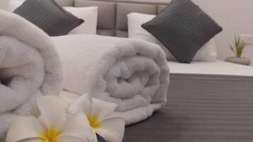 Cama arrumada com toalhas brancas dobradas, almofadas escuras, uma pequena planta em vaso e flores brancas sobre a cama.