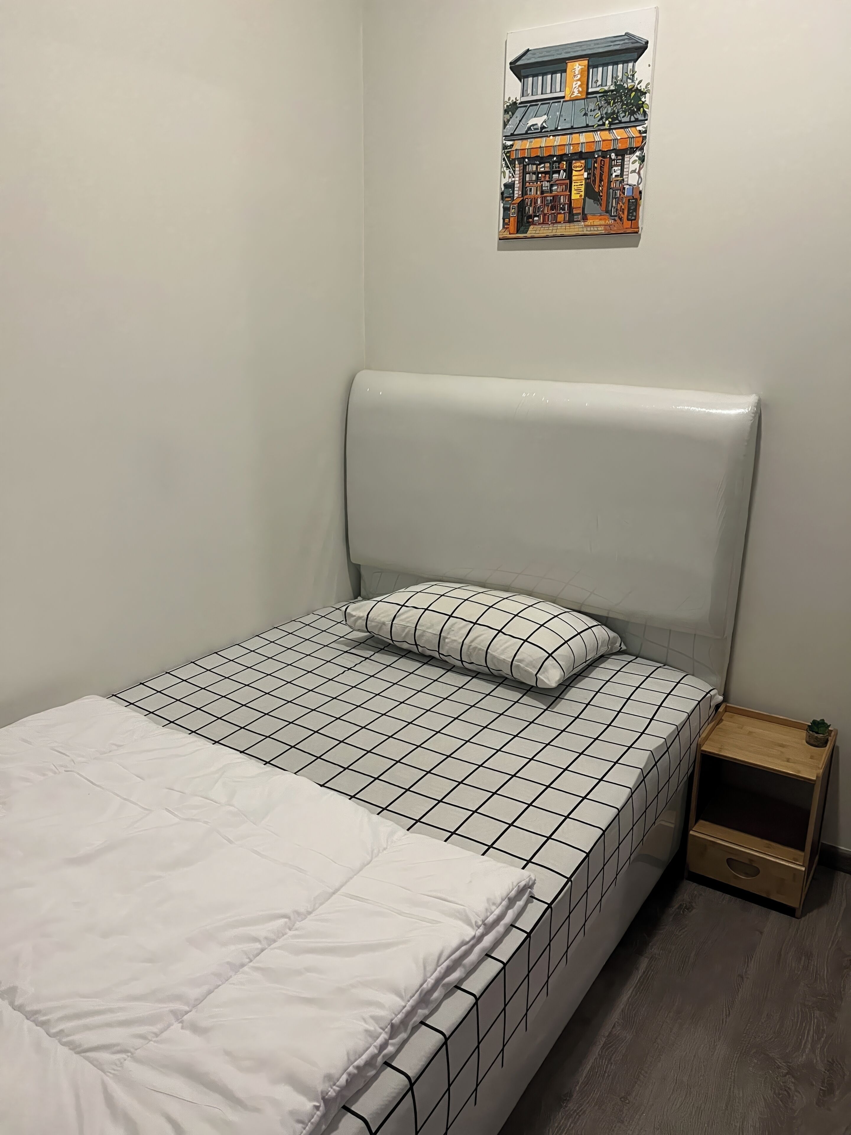 Uma cama bem arrumada com edredom quadriculado em preto e branco e almofada combinando, um pequeno criado-mudo de madeira com uma planta, e um quadro na parede com a imagem de um edifício.