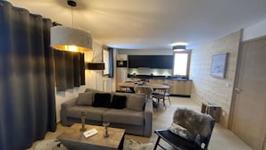 Uma sala de estar moderna com sofá, poltrona e mesa de centro. Uma cozinha é visível ao fundo, com uma área de refeições.