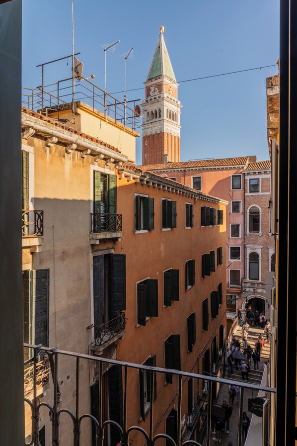 Vista de uma estreita rua veneziana, com edifícios de ambos os lados, um campanário e um céu azul límpido.