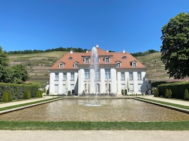 Um edifício grande e clássico com uma fonte central, rodeado por jardins bem cuidados e uma paisagem montanhosa ao fundo.
