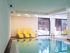 Piscina interna com água cristalina, cadeiras de descanso amarelas e vista para uma paisagem nevada através de amplas portas de vidro.