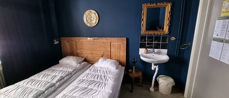 Quarto com cabeceira de madeira, duas camas com roupa de cama branca, pia branca, espelho e lixeira.
