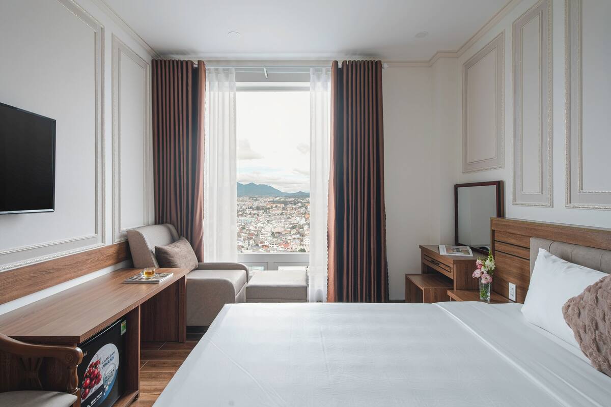 Quarto de hotel com uma cama, uma escrivaninha, uma cadeira, uma televisão e vista para a paisagem urbana pela janela.