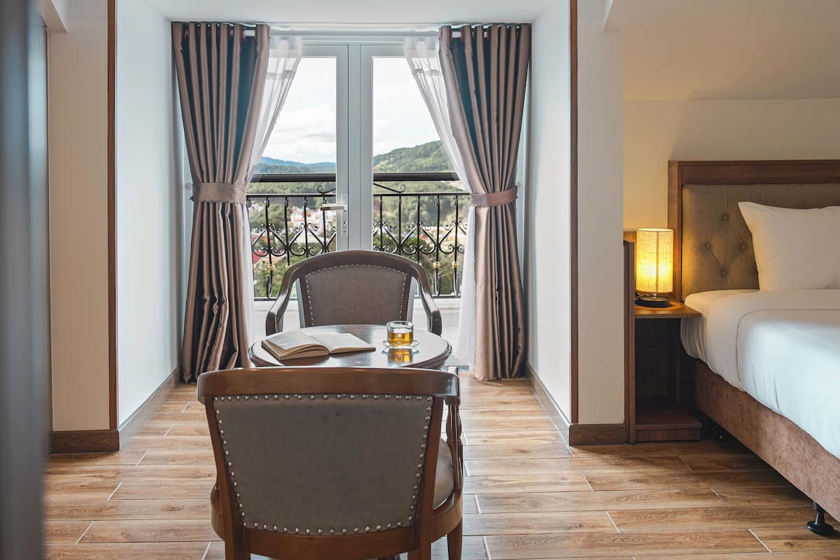 Quarto de hotel com uma cama, uma mesa com cadeira e vista para o exterior através de janelas amplas.