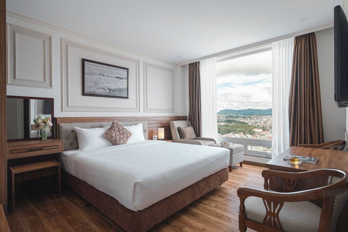 Quarto de hotel com uma cama grande, uma mesa, uma cadeira, uma televisão e uma janela com vista para a cidade.