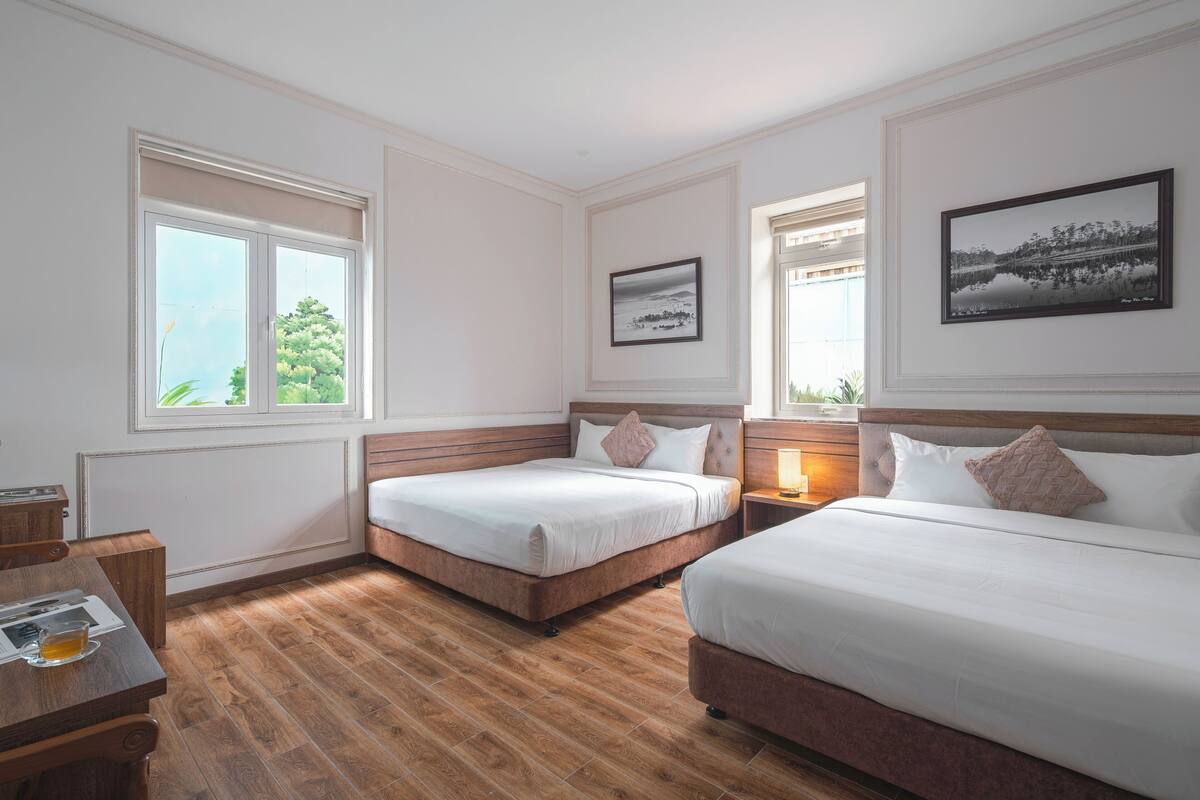 Quarto de hotel com duas camas, piso de madeira, uma escrivaninha com cadeira e uma janela com vista para o verde.