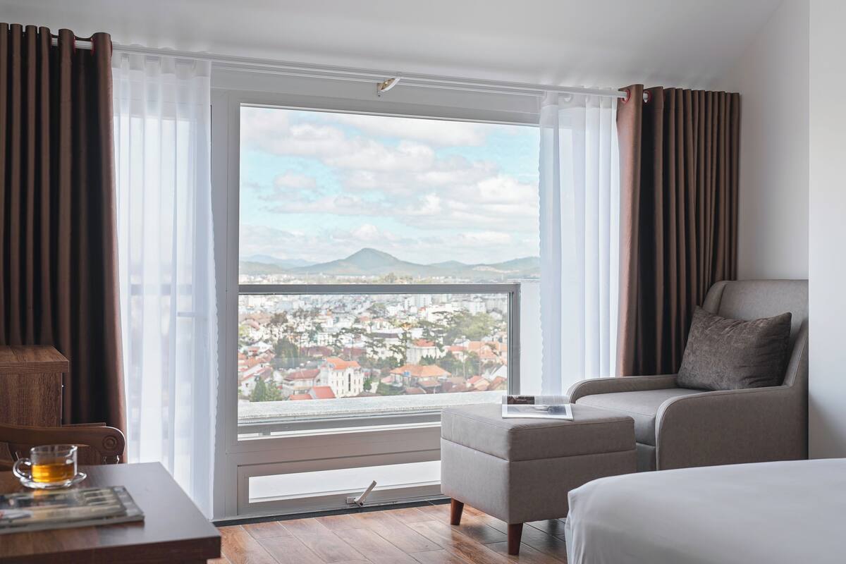 Quarto de hotel com uma janela grande, um sofá, uma mesa pequena com um copo de bebida e uma cama.