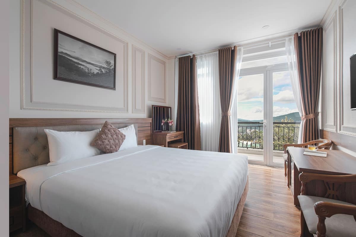 Quarto de hotel com uma cama grande, uma mesa de cabeceira de madeira, uma escrivaninha com cadeira e varanda com cortinas.