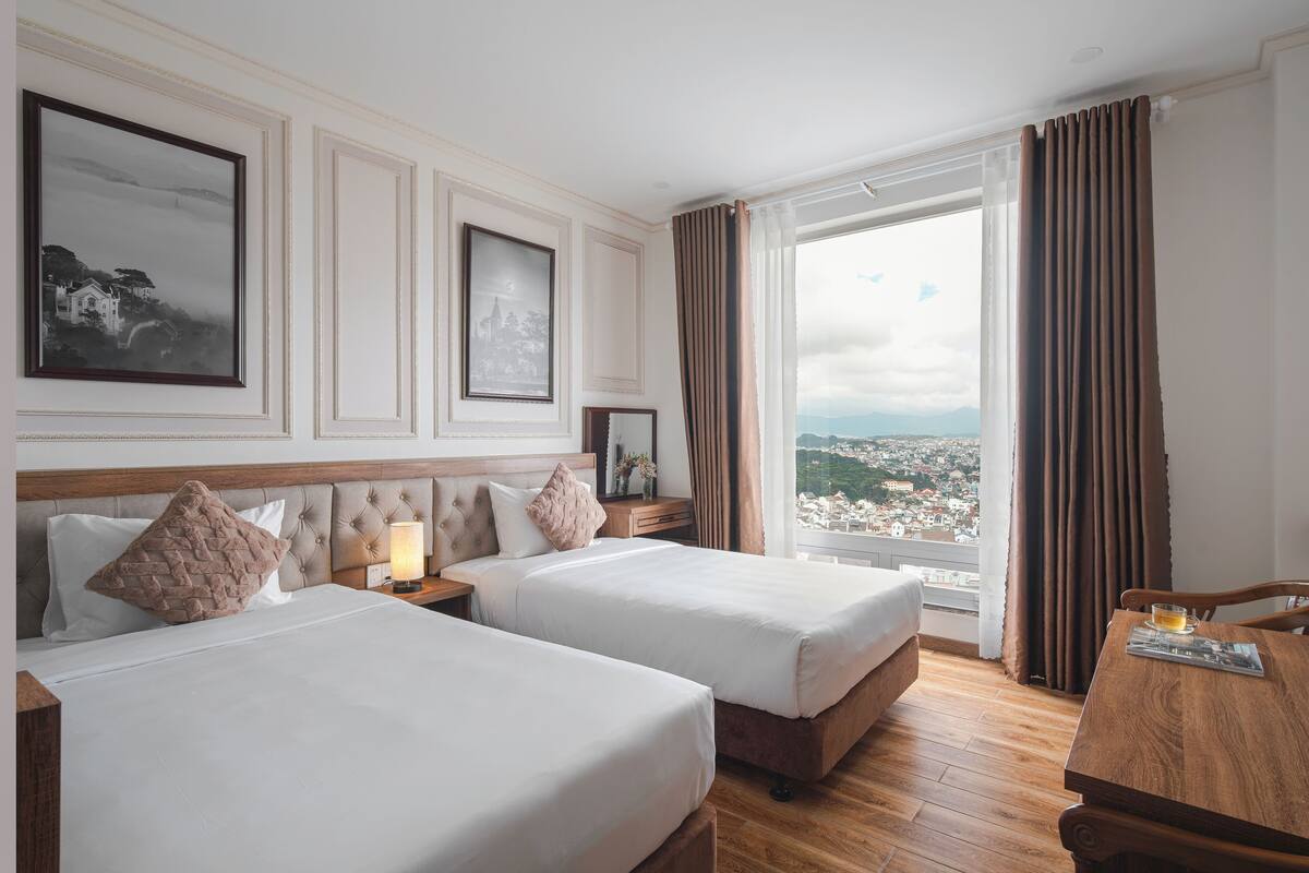 Quarto de hotel com duas camas, cabeceira de madeira, mesa de cabeceira com luminária, mesa e vista para a paisagem urbana através de janelas amplas.