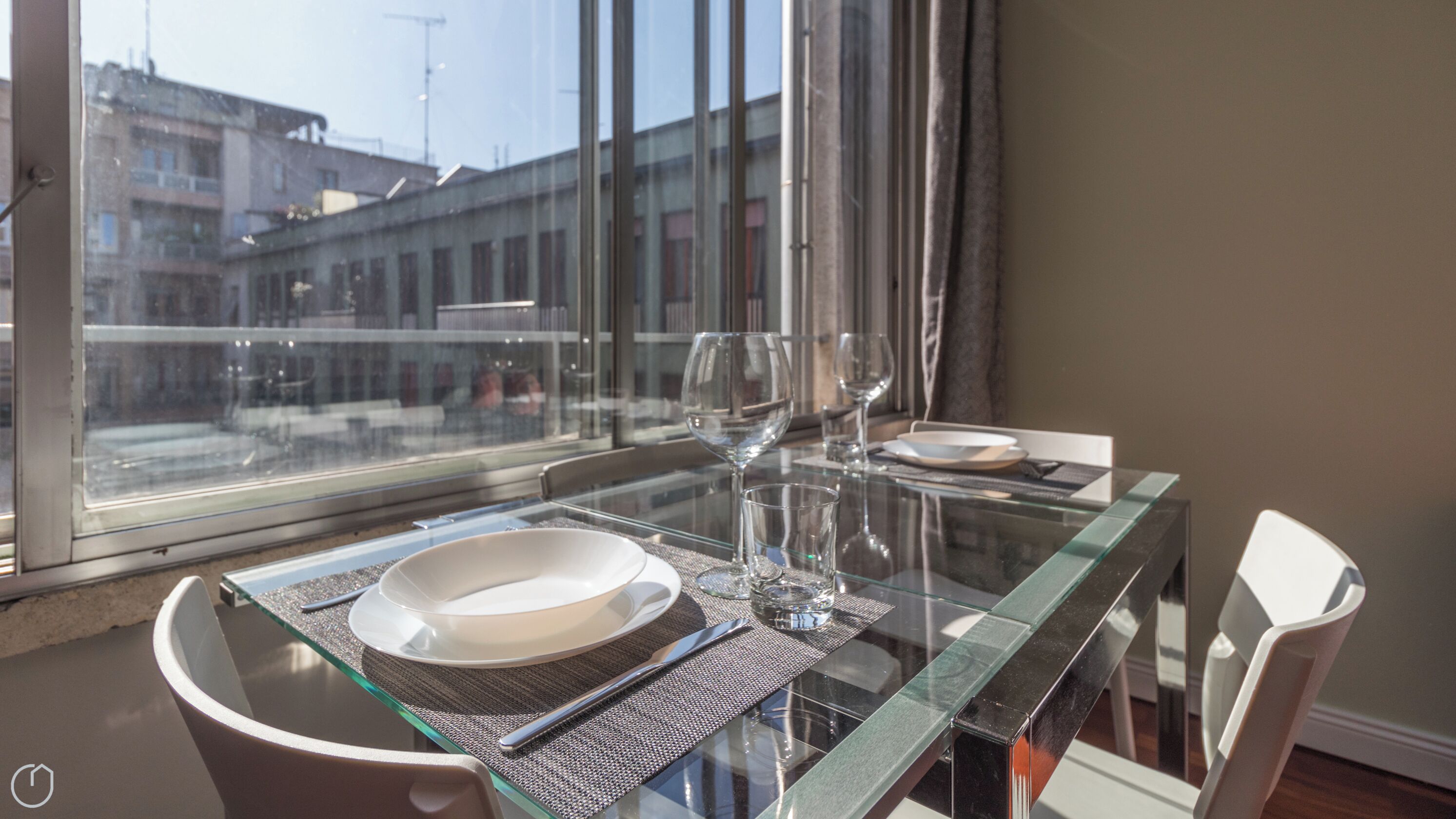 Uma mesa de jantar com pratos, copos e talheres, com vista para uma paisagem urbana através de uma janela.