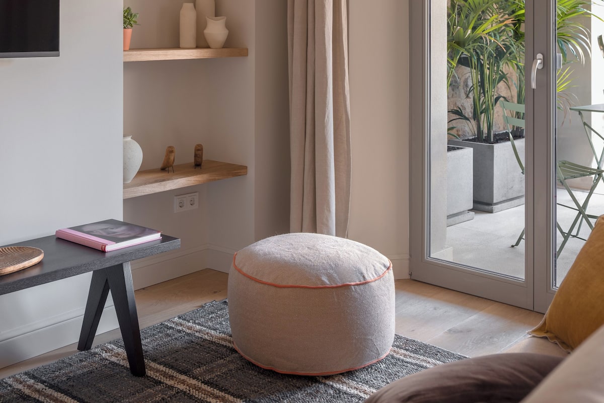 Sala de estar moderna com porta de vidro deslizante que dá para uma varanda com plantas. Há prateleiras com objetos decorativos e um banquinho redondo.
