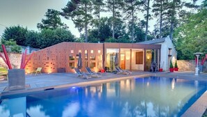 Uma área de piscina moderna ao ar livre, com um deque de madeira, espreguiçadeiras e um edifício com fachada de vidro.