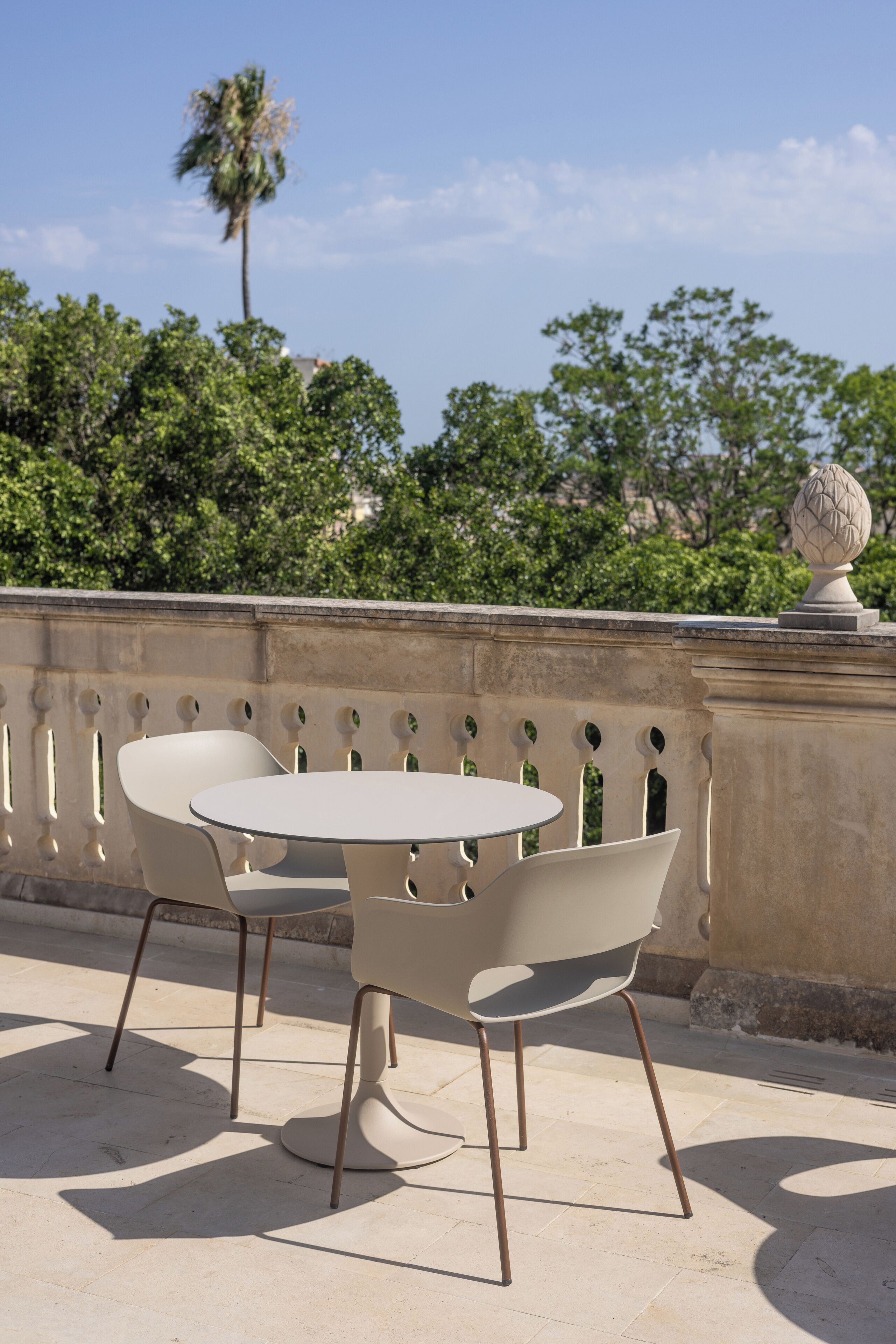 Um terraço com uma mesa redonda e duas cadeiras, um corrimão e uma palmeira ao fundo.