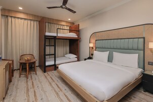Quarto de hotel com uma cama beliche, uma escrivaninha com cadeira, uma mesinha de cabeceira com abajur e um ventilador de teto.