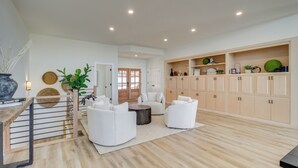 Ampla sala de estar com sofás brancos, mesa de centro e piso de madeira. Há estantes com objetos decorativos e uma escada com corrimão.