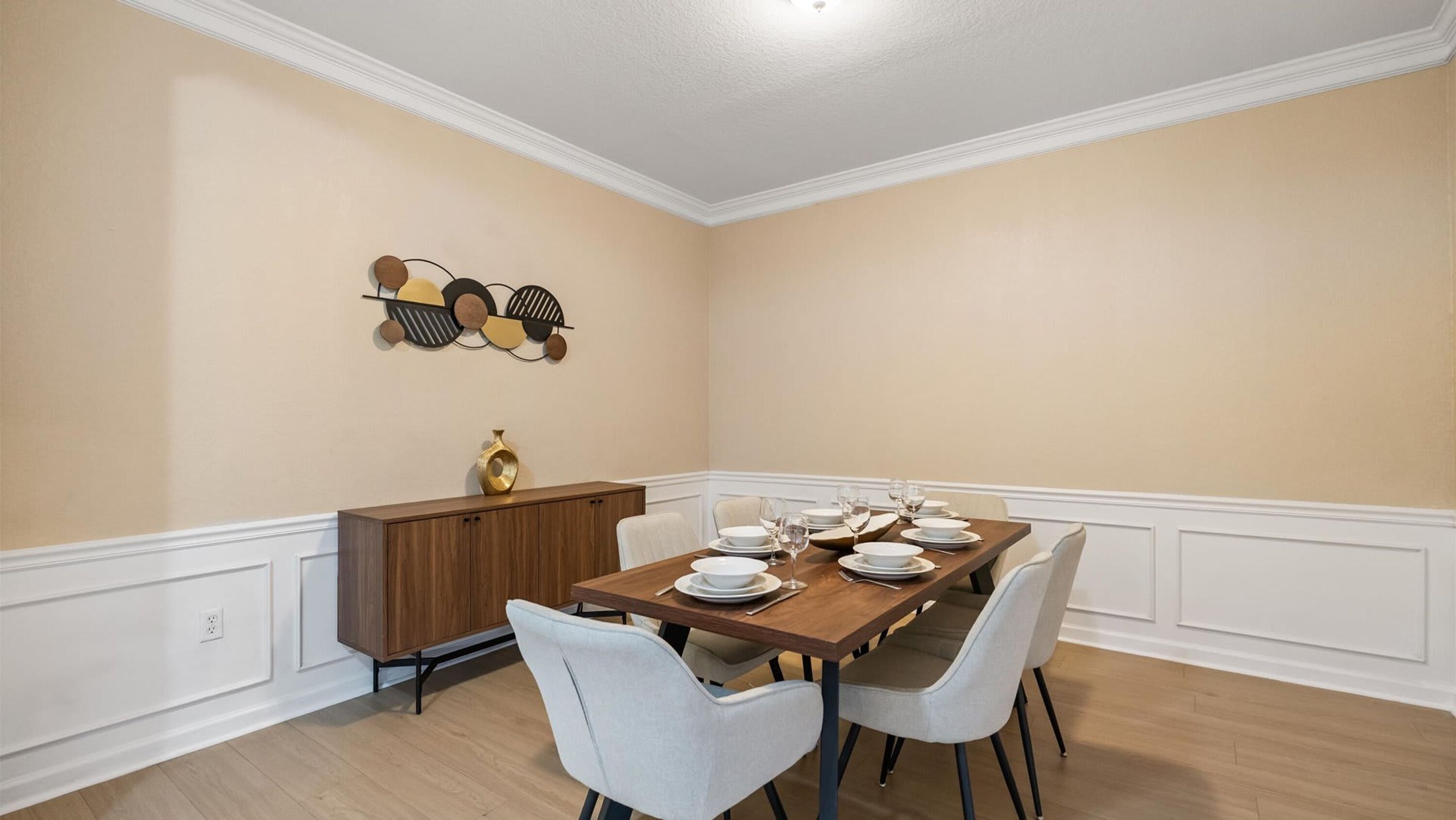 Uma área de refeições com uma mesa de madeira posta para quatro pessoas, cadeiras brancas, um detalhe decorativo na parede e um armário de madeira.