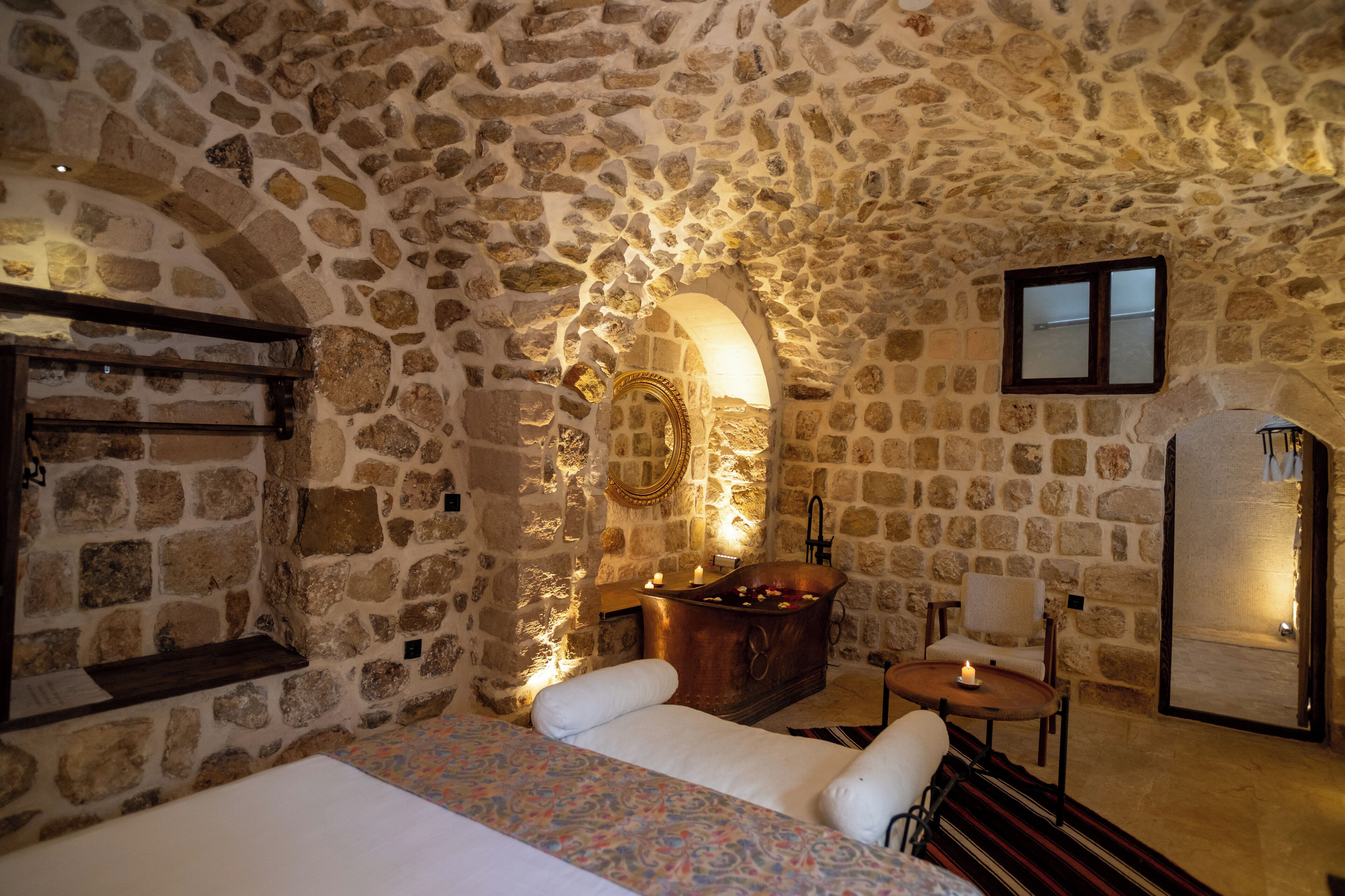 Quarto de paredes de pedra com uma cama, um banco de madeira, uma pequena mesa com uma vela e um espelho na parede.