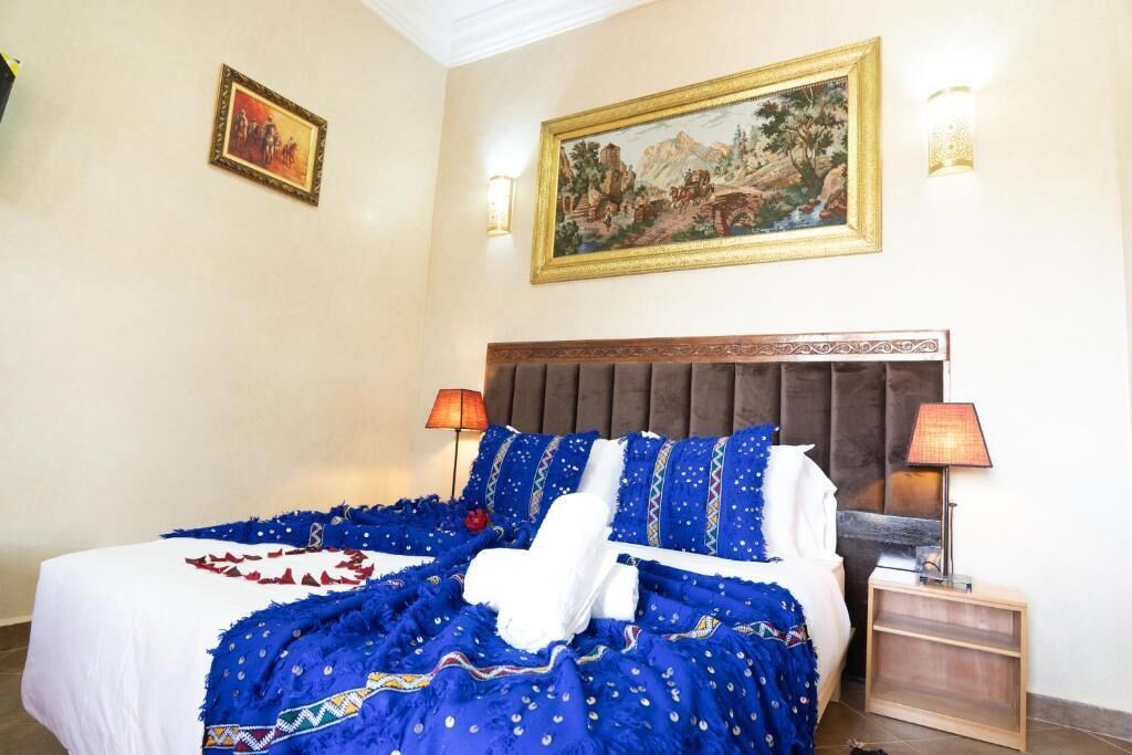 Cama com colcha estampada em azul e branco, duas almofadas e uma toalha branca. Há duas luminárias de cabeceira e uma pequena mesa de cabeceira de madeira com uma luminária.