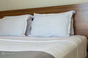 Cama bem arrumada com roupa de cama e almofadas brancas, cabeceira de madeira e o logotipo de Andrade Hotels.