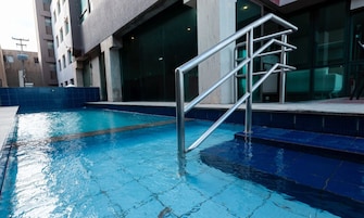Piscina com escada de metal para acesso à água, rodeada por um deck ladrilhado e um edifício com grandes janelas.