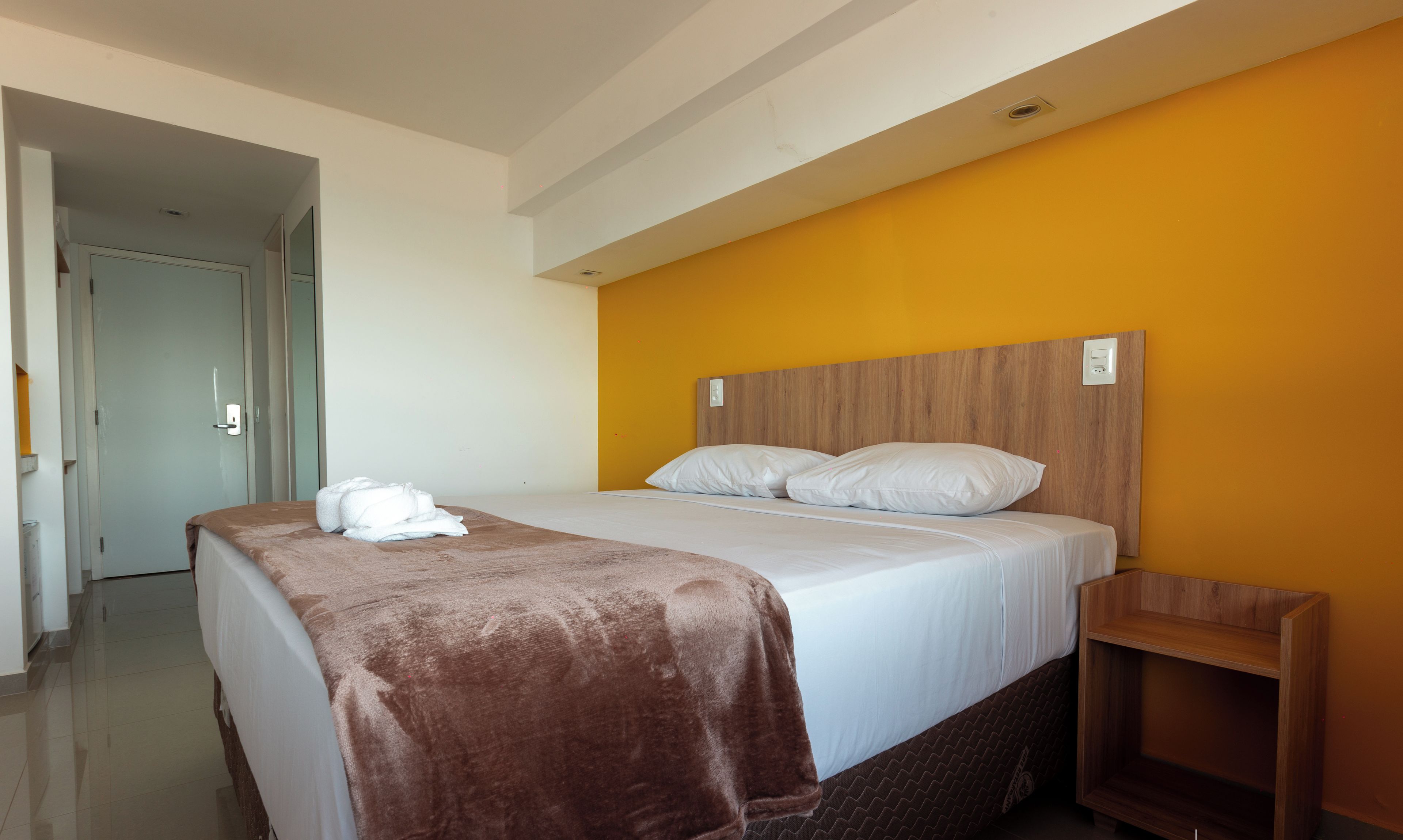 Quarto de hotel com cama, cabeceira de madeira, manta marrom e toalha branca.