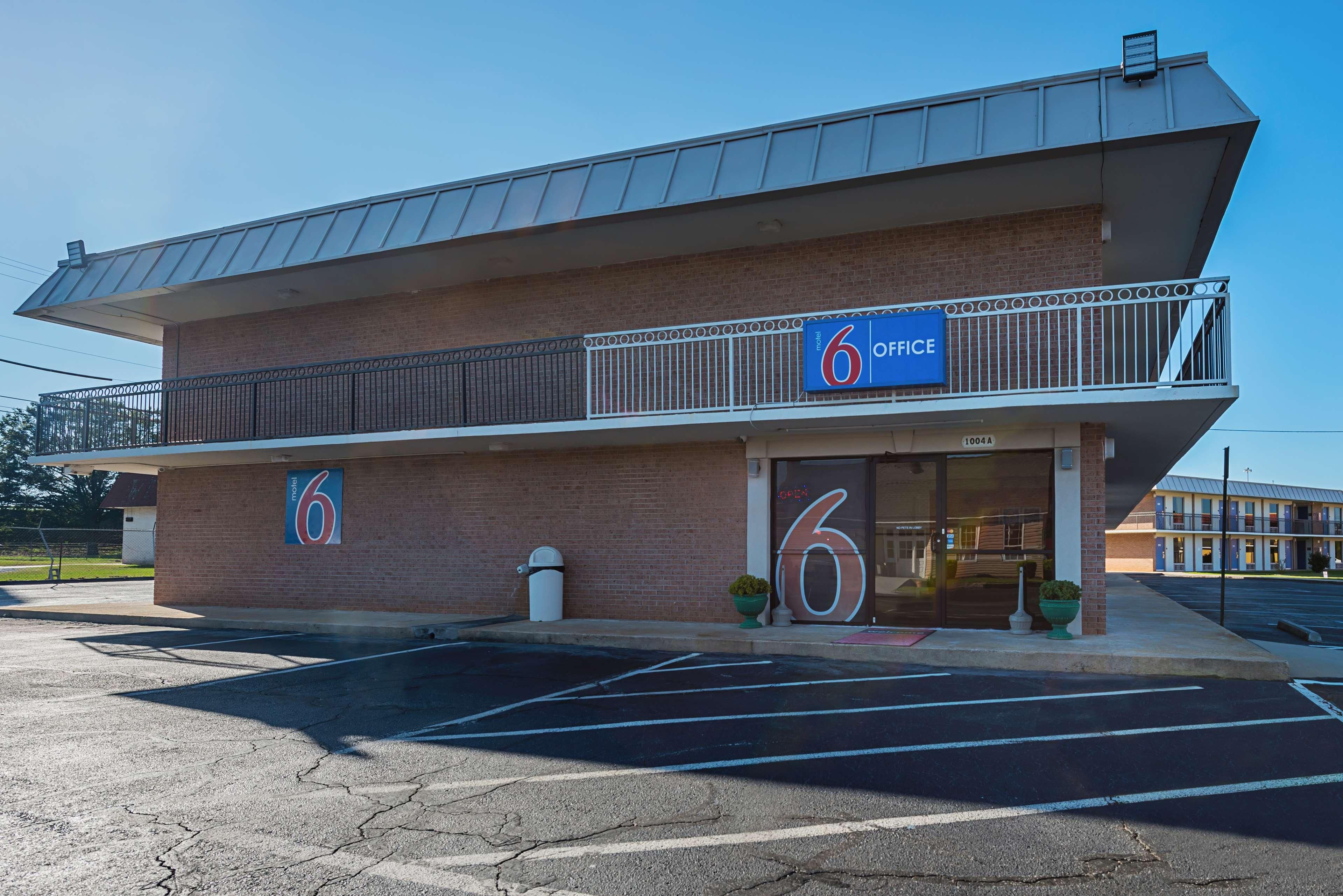 Um motel de dois andares com uma placa grande '6 Office', um estacionamento e fachada de tijolos.