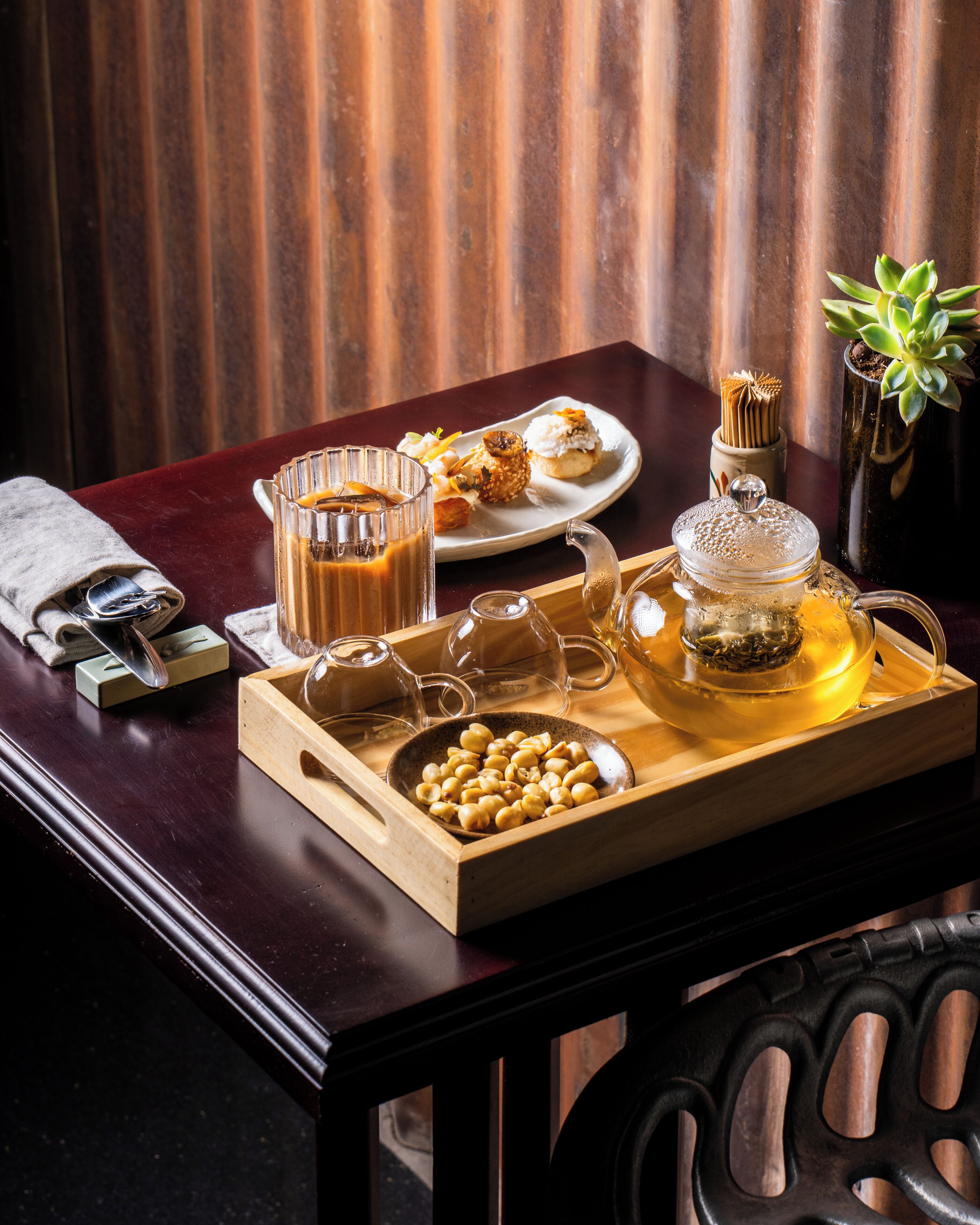 Uma bandeja de madeira com uma chávena de café, uma taça de frutos secos e uma chaleira sobre uma mesa.