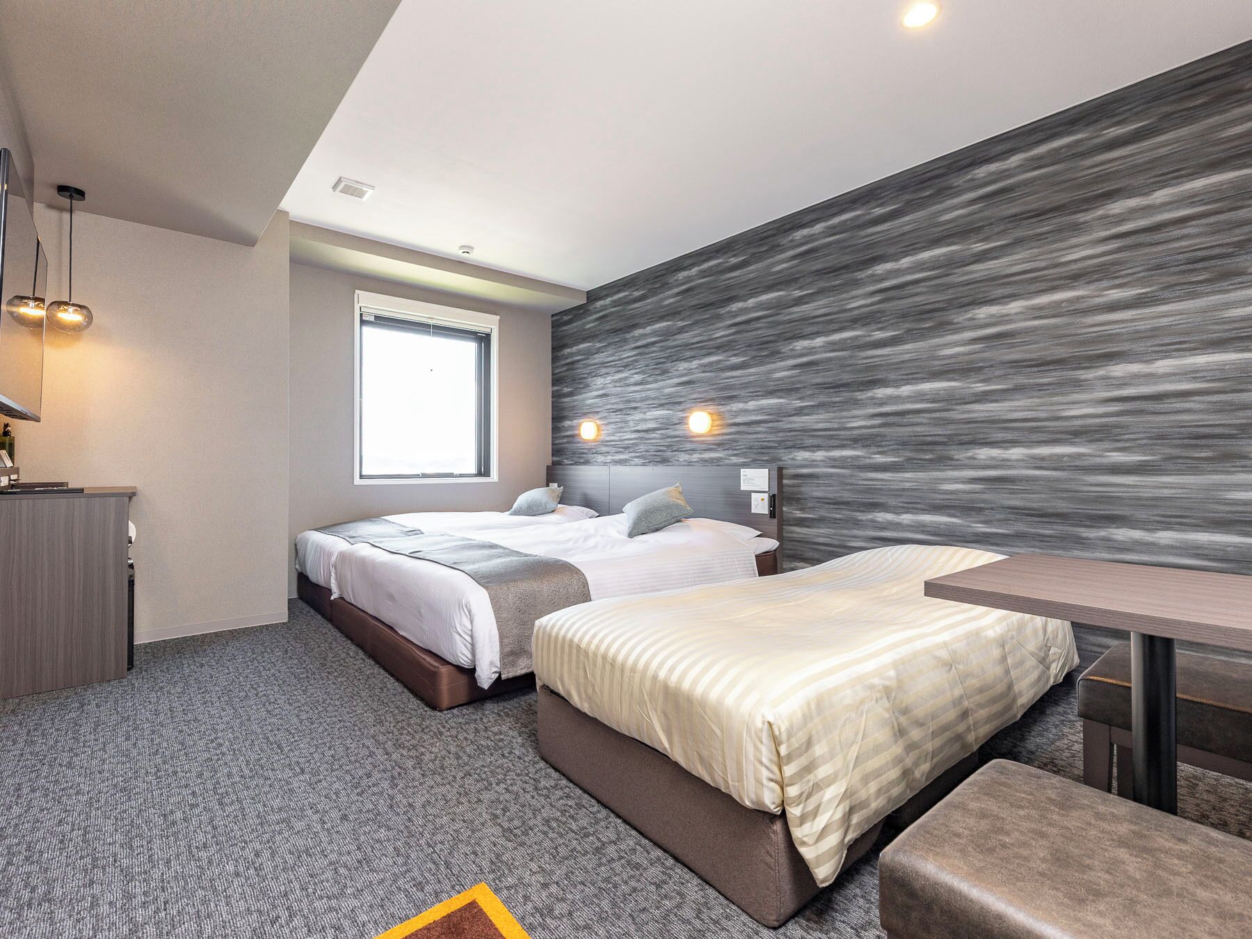 Quarto de hotel moderno com duas camas, uma escrivaninha e uma janela grande.