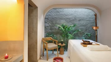 Um quarto de spa com uma cadeira de madeira, uma mesinha com um vaso, um recipiente com flores e uma grande janela em arco com vista para vegetação.