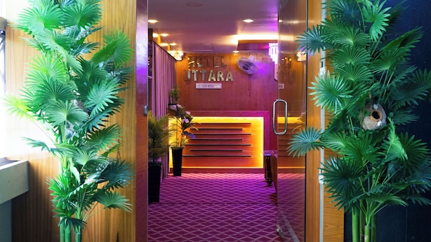 Uma área de recepção de hotel com um balcão de madeira, placa de recepção e plantas em vasos.