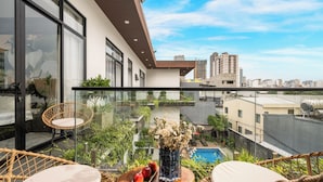 Varanda com cadeiras de vime, mesa de madeira com frutas e flores, e vista para a cidade e uma piscina.