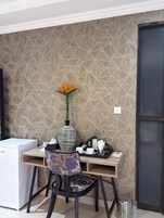Quarto de hotel moderno com parede estampada em padrão geométrico, área de refeições com refrigerador branco, vaso com padrão preto e branco e cadeira azul com estampa floral.