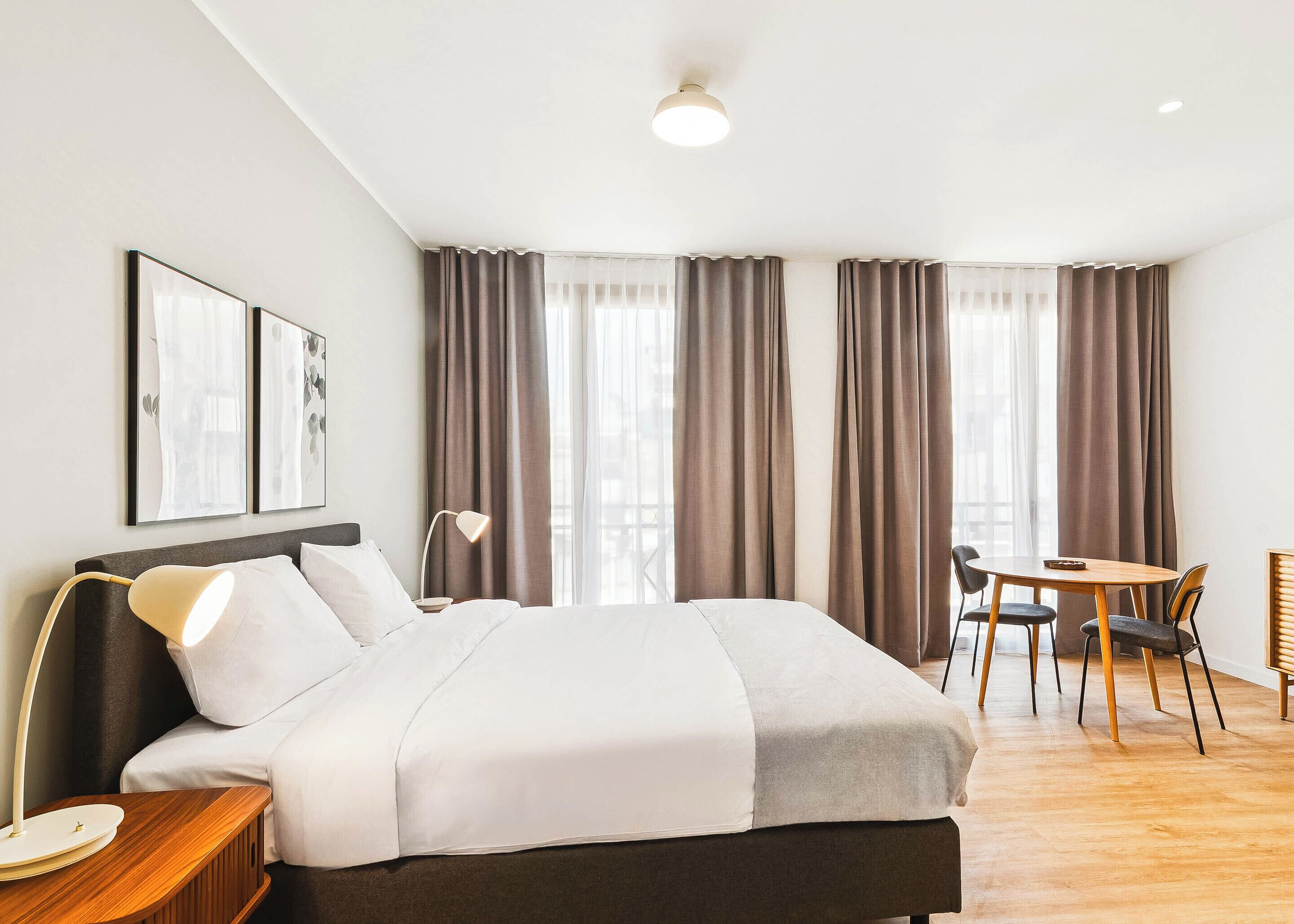 Quarto de hotel moderno com uma cama grande, mesa de cabeceira com abajur, pequena mesa com cadeiras e janela com cortinas.