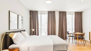 Quarto de hotel moderno com uma cama grande, mesa de cabeceira com abajur, pequena mesa com cadeiras e janela com cortinas.