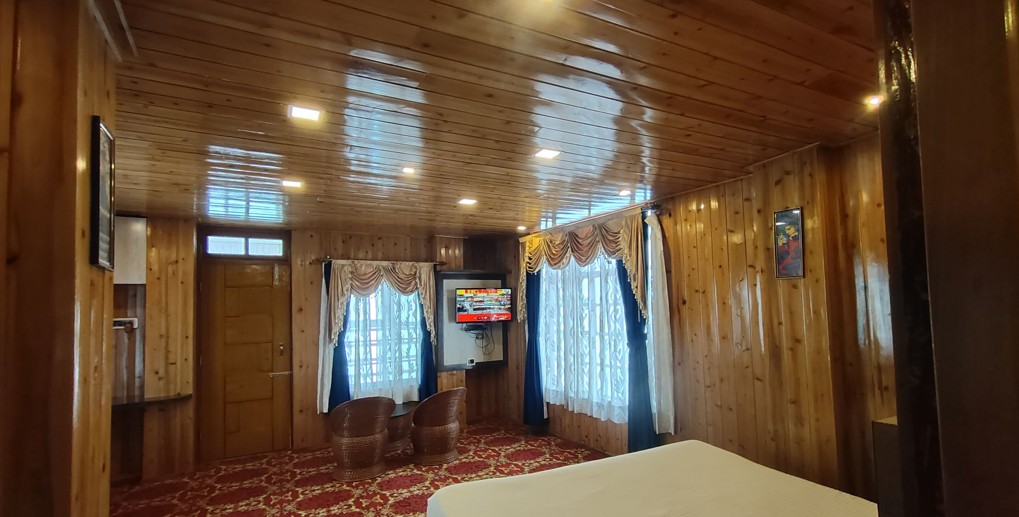 Um quarto com painéis de madeira, uma cama, duas cadeiras de vime, uma televisão e um tapete vermelho.