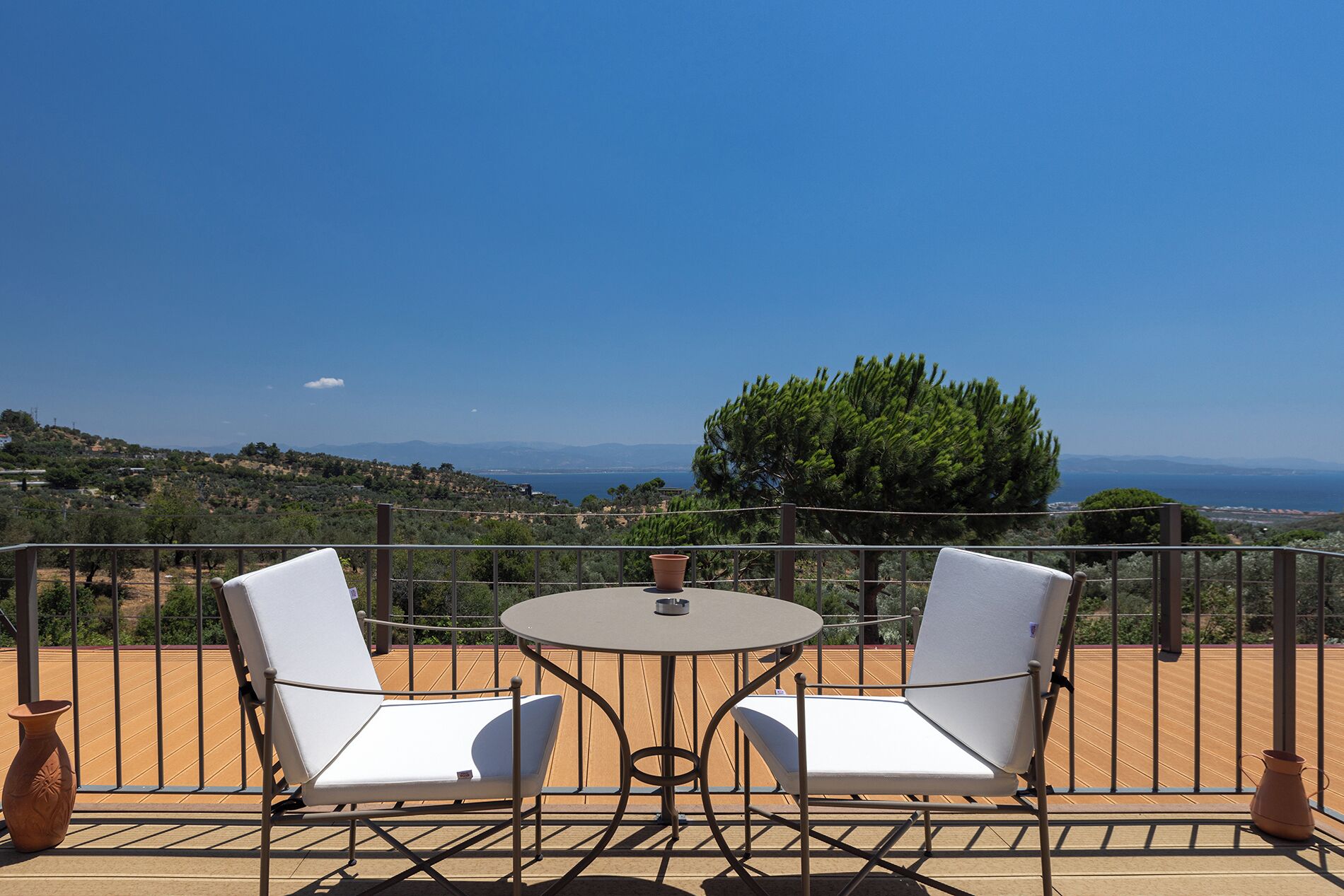 Terraço com duas cadeiras brancas e uma mesa, com vista para uma paisagem pitoresca com colinas e o mar.