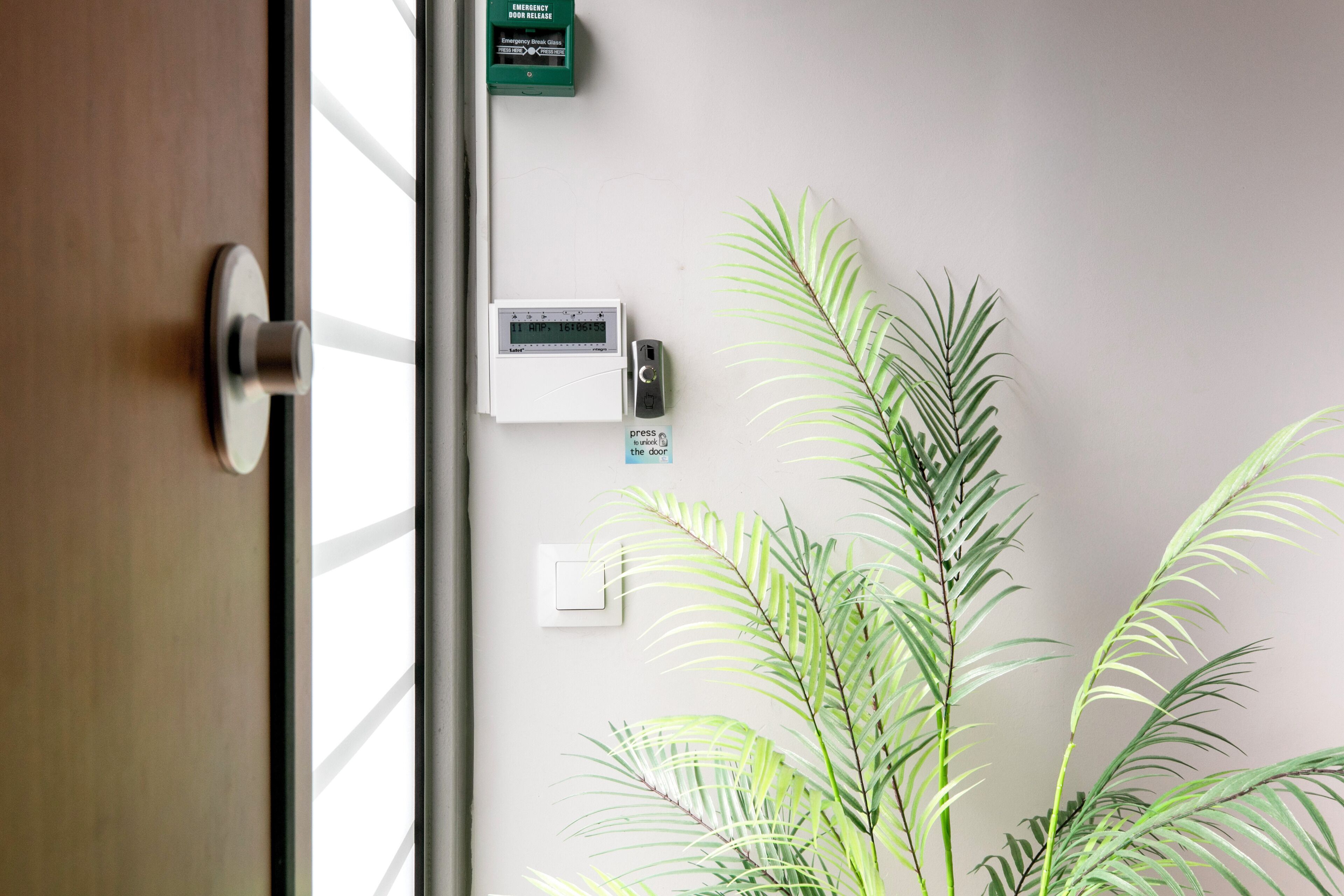 Uma porta com fechadura digital, um dispositivo de liberação de emergência fixado na parede, um interruptor de luz e um vaso de planta.