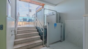 Escadaria que leva a um patamar com porta de vidro, sinal de acessibilidade para cadeiras de rodas e vista para área de estar externa.