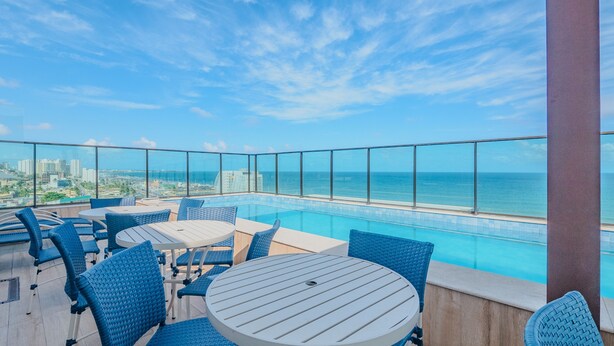 Uma área de piscina no terraço, com cadeiras e mesas azuis, com vista para a paisagem urbana e o oceano.