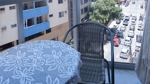 Uma varanda com mesa e cadeira, dando para uma rua com carros estacionados e edifícios altos ao fundo.