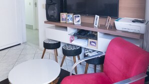 Uma sala de estar com televisão, uma cadeira vermelha, uma mesa branca e um sofá pequeno.