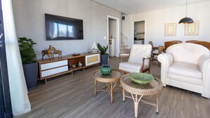 Uma sala de estar moderna com televisão de tela plana, um sofá, duas mesinhas de vime e uma poltrona branca.