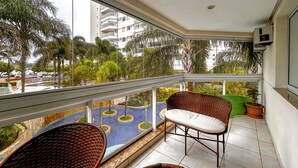 Uma varanda com cadeiras de vime, uma mesa verde e vista para um jardim com palmeiras e um edifício ao fundo.