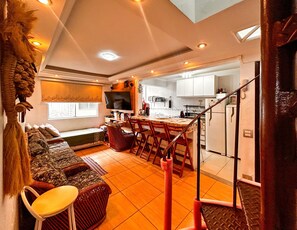 Uma sala de estar acolhedora com sofá, cadeiras e uma televisão. Uma cozinha é visível ao fundo, com armários e eletrodomésticos.