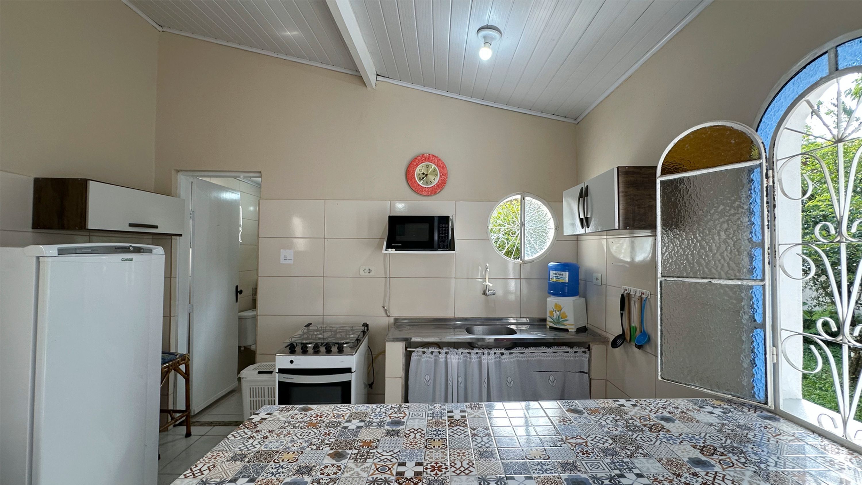 Cozinha compacta com geladeira, fogão, micro-ondas e pia. Há uma janela com portão de metal decorativo e um relógio na parede.
