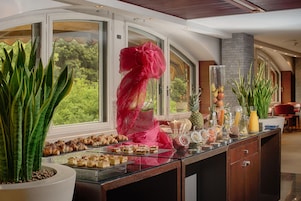 Uma área de buffet com diversos tipos de comida, uma grande planta em vaso e um laço rosa decorativo.