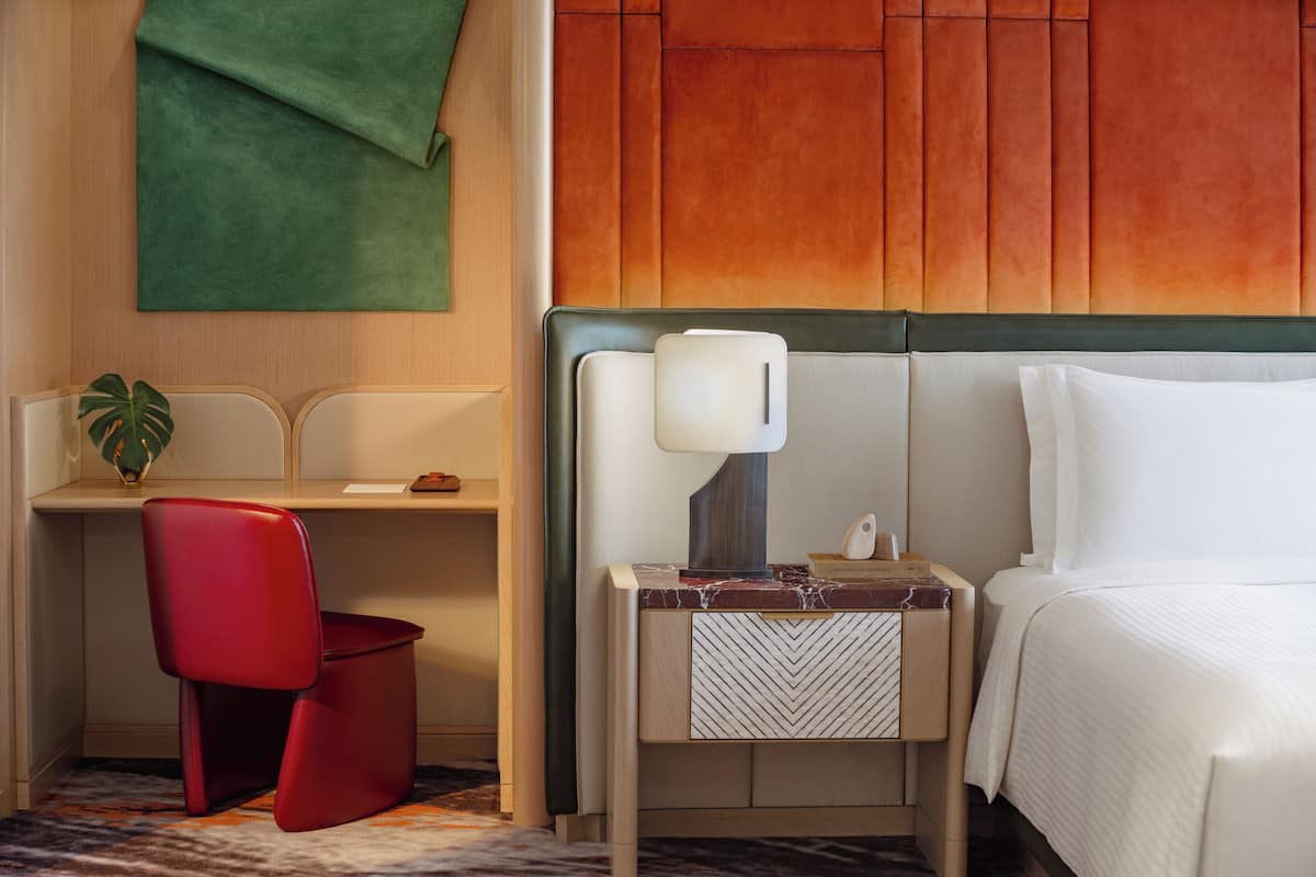 Quarto de hotel com cama, escrivaninha com cadeira, mesa de cabeceira com abajur e um painel verde na parede.