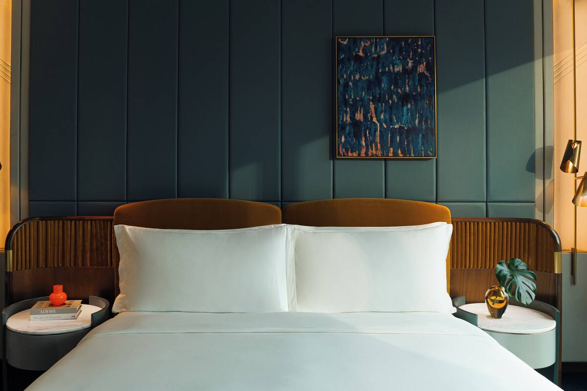 Quarto de hotel moderno com uma cama grande, duas mesas de cabeceira, um criado-mudo, uma planta em vaso e um quadro na parede.