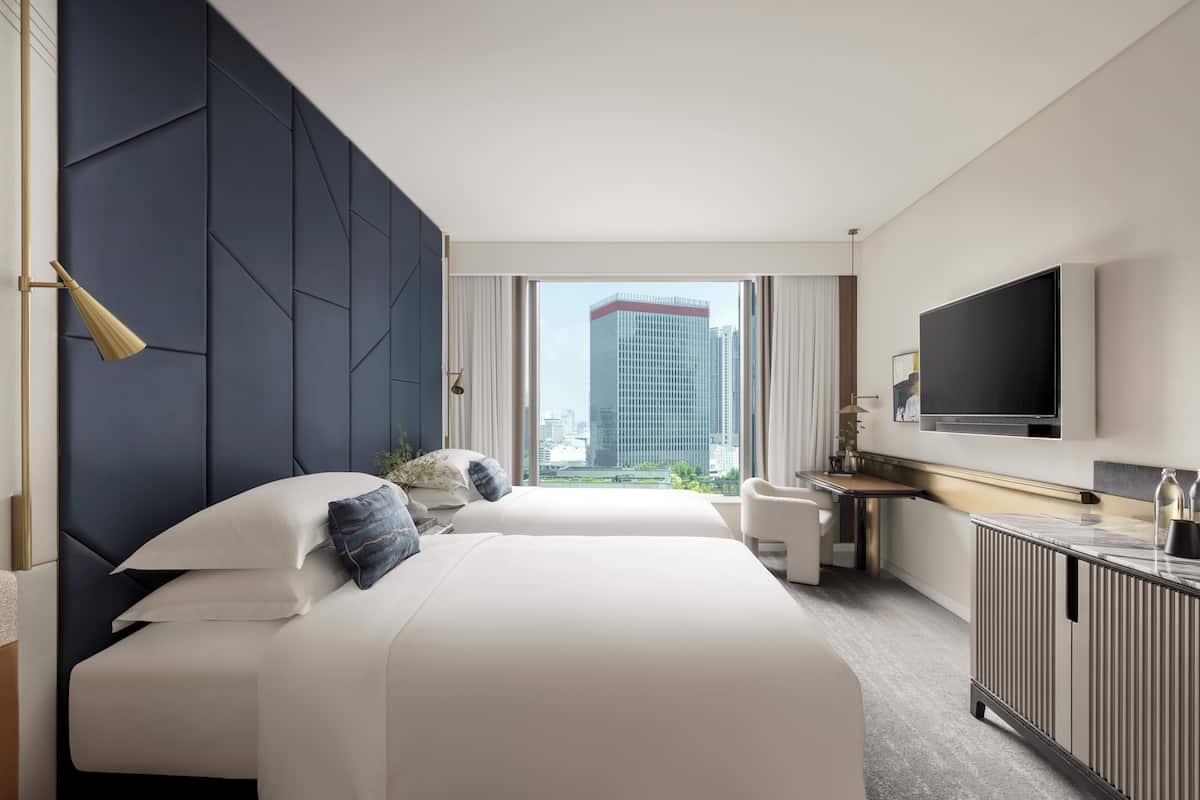 Quarto de hotel moderno com uma cama grande, uma mesa de trabalho, televisão e vista para a paisagem urbana.